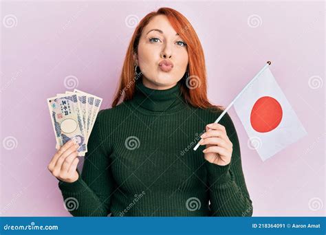 Beautiful Redhead Woman Holding Japanese Yen Banknotes And Japan Flag Looking At The Camera