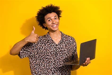 Chevelure 착용 표범 셔츠를 가진 낙관적 인 재미 있는 사람의 초상화는 노란색 배경에 고립 된 노트북 쇼 엄지 손가락을 보여줍니다 갈색 머리에 대한 스톡 사진 및