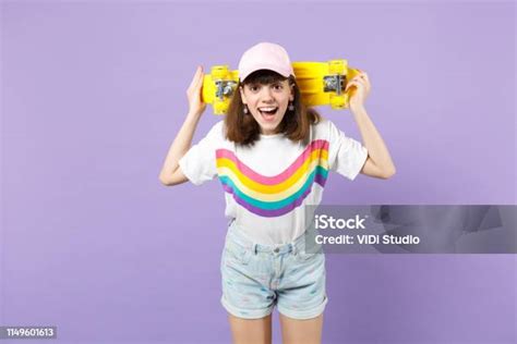 스튜디오에서 보라색 파스텔 벽 배경에 고립 된 노란 스케이트 보드를 들고 생생한 옷에 흥분 웃는 십 대 소녀의 초상화 사람들은