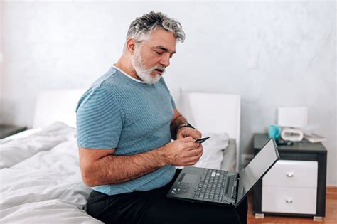 침대에서 쇼핑 중년 남성은 노트북과 신용 카드를 손에 들고 온라인 거래를 걸었습니다 50 54세에 대한 스톡 사진 및 기타