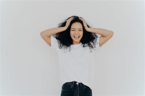 기뻐하는 곱슬 머리 의 여자 행복하게 웃고 흰색 티셔츠와 청바지를 입고 흰색 배경위에 고립 된 광범위하게 미소를 짓고 있습니다