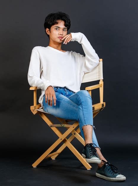 검은 배경에 몸짓으로 포즈를 취하는 나무 의자에 다리를 꼬고 앉아 캐주얼 복장을 한 아시아 젊은 Lgbtq 게이 글래머 잘생긴 양성애자 동성애 남성 모델의 전신 사진