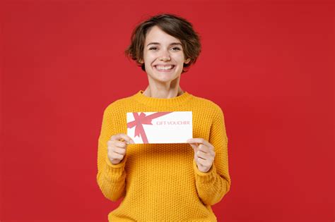 밝은 빨간 색 벽 배경 스튜디오 초상화에 고립 된 손 상품권에 들고 캐주얼 노란색 스웨터를 입고 웃는 쾌활한 매력적인 젊은 갈색