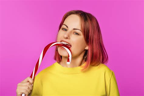 행복한 예쁜 젊은 여자가 달콤한 사탕을 핥고 롤리팝 룩 카메라를 먹는다 분홍색 머리와 분홍색 배경 위에 서 있는 노란색 티셔츠를 입은 사랑스러운 여성 모델 30 34세에