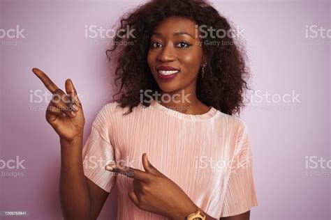 고립된 분홍색 배경 위에 서서 미소를 짓고 두 손과 손가락으로 측면을 가리키는 카메라를 바라보는 젊은 아프리카 계 미국인 여성 T 셔츠에 대한 스톡 사진 및 기타 이미지
