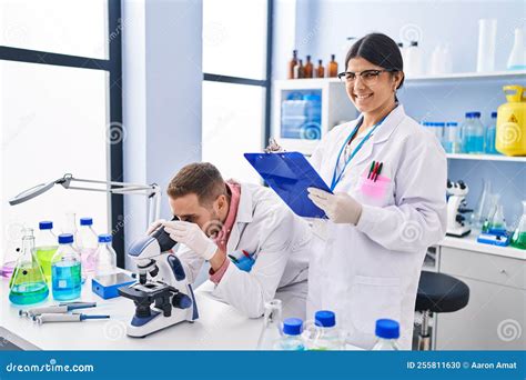 Man And Woman Wearing Scientists Uniform Using Microscope At Laboratory