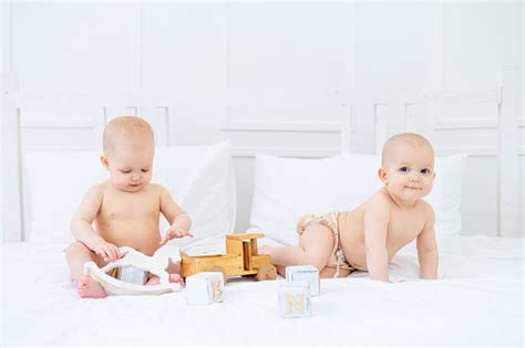 두 쌍둥이 아기 6개월 동안 기저귀를 찬 소녀들이 흰색 면 침대에 바디수트를 입고 집에서 나무 장난감을 가지고 놀고 웃고 있다