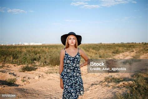 꽃 무늬와 일몰 동안 여름에 포도 밭 근처 모래 마을 도로에 서 검은 모자와 파란색 드레스에 젊은 아름다운 백인 금발 소녀의 초상화 가냘픈에 대한 스톡 사진 및 기타 이미지