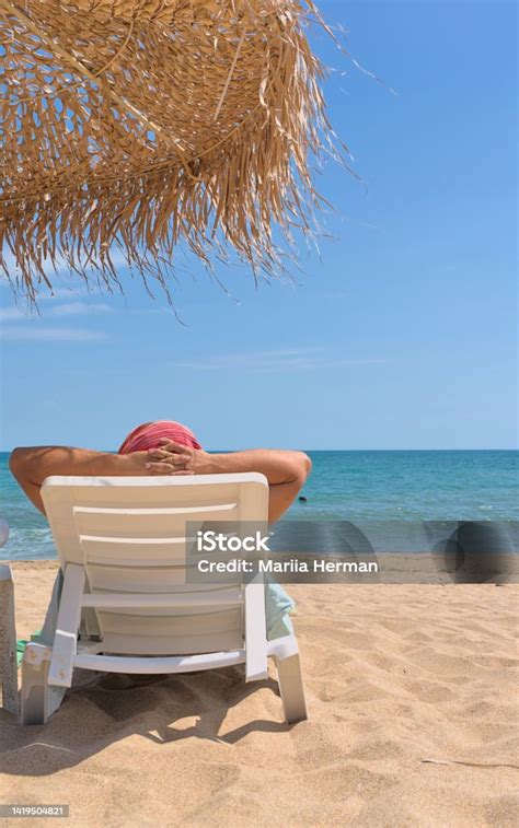 한 남자가 짚 우산 아래 바다 근처의 모래 사장에있는 하얀 일광욕 용 의자에 누워 있습니다 파도와 안개로부터 보호되는 스노클링 마스크가 일광욕 용 의자에 매달려 있습니다