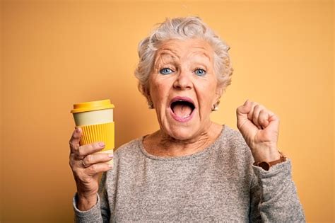 고립 된 노란색 배경 위에 서있는 커피 잔을 마시는 선임 아름다운 여성은 자랑스럽게 비명을 지르며 승리와 성공을 축하하며 매우 흥분하고 응원하는 감정을 감정에 대한 스톡