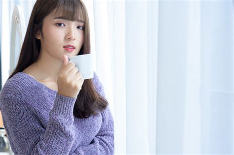 아시아 여자는 창에서 밝은 배경에서 그녀의 손에 커피 한 잔 들고 그녀는 스웨터 바이올렛 컬러 드레스 모닝 커피 시간입니다