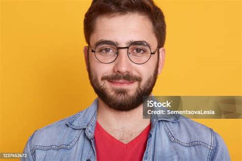 카메라를 직접 바라보며 진지하게 웃고 안경 데님 재킷 빨간 티셔츠를 입고 기분 좋게 웃고 있는 유쾌한 젊은 남자의 실내 수평 이미지 감정 개념 귀여운에 대한 스톡 사진 및