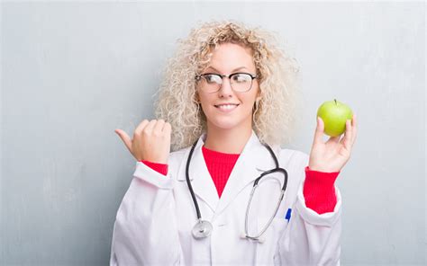 젊은 금발 영양사 의사 여자 고 행복 얼굴 미소와 측면까지 손가락으로 보여주는 녹색 사과 들고 그런 지 회색 벽에 가리키기에 대한 스톡 사진 및 기타 이미지 Istock