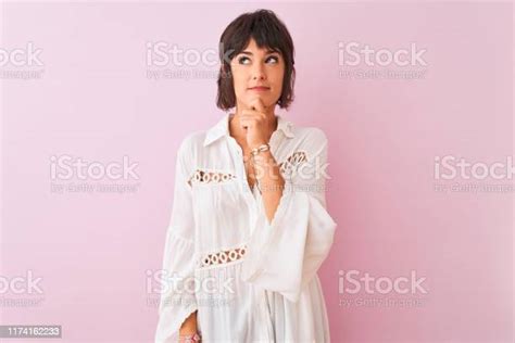 여름 흰색 셔츠를 입고 있는 젊은 아름 다운 여자는 고립 된 분홍색 배경 위에 서 턱에 손을 대 고 질문 펜시브 표정에 대 한