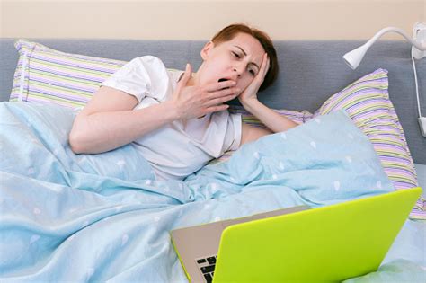 여자는 덮개 아래 침대에 누워 노트북에 영화를 보고 여자 하품 커버 그녀의 입 와 그녀의 손 35 39세에 대한 스톡 사진 및