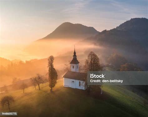 가을의 황금빛 일출에 낮은 구름 속에서 언덕에 있는 작은 교회의 공중 전망 슬로베니아 아름다운 예배당 산골 마을 안개 푸른 초원 나무 가을 새벽 하늘의 최고 전망 풍경화