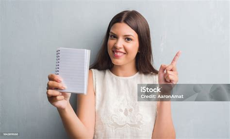 그런 지 위에 젊은 갈색 머리 여자 회색 벽 보여주는 노트북 아이디어 놀된 또는 행복 한 얼굴 넘버 원 가리키는 손가락 질문