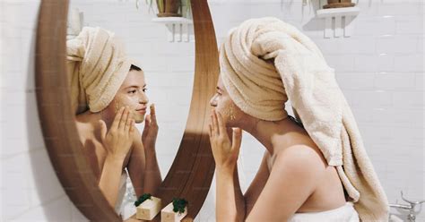 수건을 쓴 젊은 행복한 여자는 유기농 얼굴 마스크를 쓰고 세련된 욕실에서 둥근 거울을 보고 있다 얼굴의 피부를 스크럽하고 껍질을 벗기고 청소하는 얼굴 마사지를 만드는 소녀