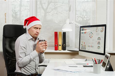 잘생긴 심각한 비즈니스 남자 셔츠와 책상에 앉아 빨간 크리스마스 모자에 차 또는 커피 한잔으로 몸을 따뜻하게 빛 사무실에서 문서와 현대 모니터 컴퓨터에서 작업 경영자에 대한