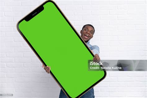 흥분한 흑인 남자가 서서 빈 녹색 화면이있는 큰 스마트 폰을 가리키고 앱 또는 웹 사이트 디자인을위한 복사 공간을 시연하고 흰색 벽돌 배경 위에 서서 모형 이미지를 보여
