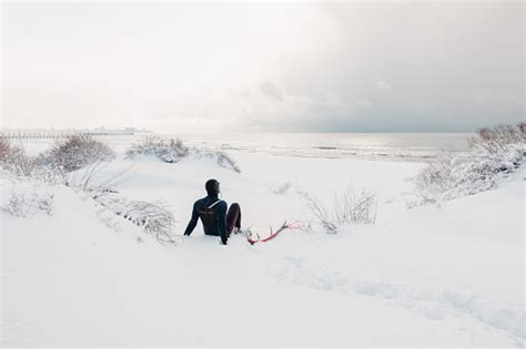 추운 겨울과 서퍼는 서핑 보드와 눈 덮인 해변에 앉아 파도에서 볼 수 있습니다 잠수복을 입은 서퍼와 함께 하는 겨울날 눈 냉동