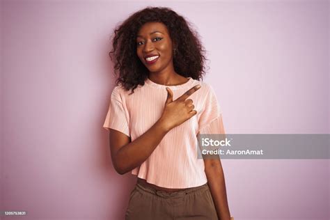 고립된 분홍색 배경 위에 서있는 티셔츠를 입은 젊은 아프리카 계 미국인 여성은 얼굴에 행복하고 자연스러운 표정으로 손과 손가락으로 가리키는 얼굴의 미소와 함께 명랑합니다 T