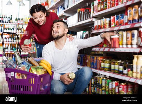 Happy Cheerful Couple Standing Near Shelves With Canned Goods At Store