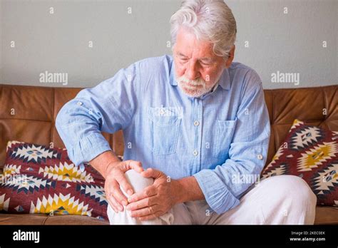 Senior Old Man Facing Knee Problem Sitting On Sofa Holding Knee At