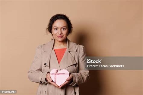 다민족 예쁜 여자는 카메라를 보고 미소를 짓고 귀여운 분홍색 하트 모양의 선물 상자 고립된 베이지색 배경을 들고 있다 갈색 머리에 대한 스톡 사진 및 기타 이미지 Istock