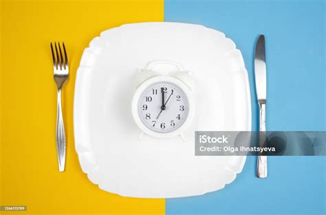 흰색 알람 시계는 파란색 노란색 배경 상단 보기에 측면에 포크와 칼흰색 접시에 놓여 0명에 대한 스톡 사진 및 기타 이미지