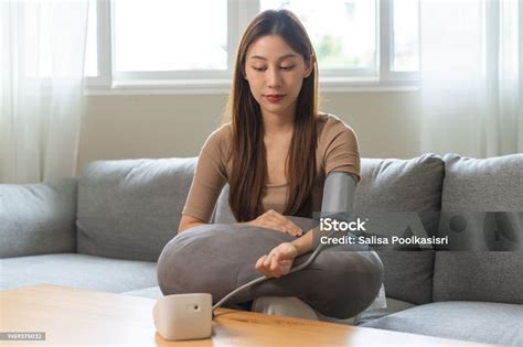 저혈압 소파에 앉아 디지털 압력 모니터 기계로 혈액 고압과 심박수를 확인하는 아시아 젊은 여성이 집에서 팔을 자가 검진합니다