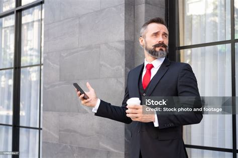 얼굴에 선택적 초점 수염을 기고한 성인 백인 사업가 중앙 비즈니스 지구에 서 있는 정장 차림 아침 커피를 마시고 자신의 모바일 스마트 폰을 진지하게 보는 동안 남자 사업가에