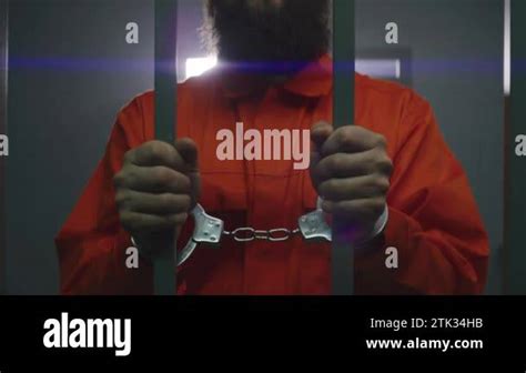 Male Criminal In Orange Uniform And Handcuffs Holds Metal Bars Stands