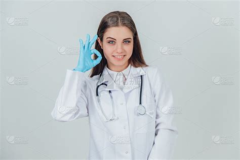受鼓舞的年轻女医生站着，穿着医疗制服，对着镜头微笑，显示ok的迹象。