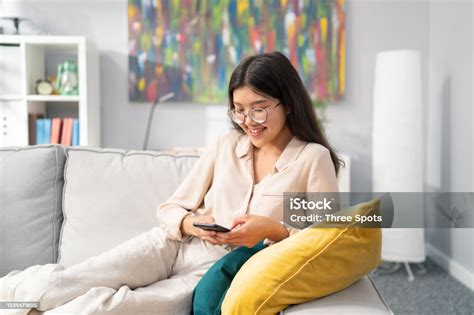 독특한 이국적인 아름다움을 가진 어린 예쁜 소녀는 다리를 침대를 따라 펼쳐진 방에있는 소파에 앉아 전화로 손가락을 클릭하고 메시지를 보내고 게시물에 댓글을 달고 친구에게