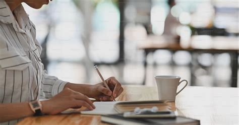 흰색 줄무늬 셔츠를 입은 젊은 비서 여성이 나무 책상에 앉아 노트북에 글을 쓰거나 메모를 하는 장면 사진 Unsplash의 컵 이미지