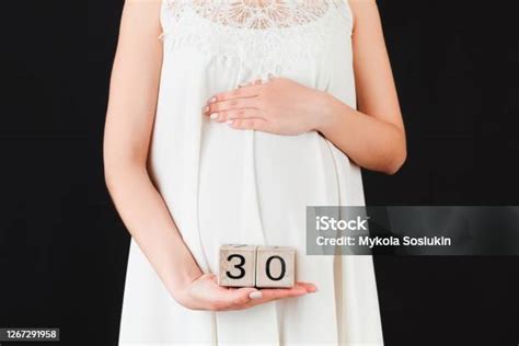 검은 색 배경에서 임신 큐브의 30 주를 보여주는 흰색 드레스에 임신 한 여성의 자른 이미지 아기를 기다리고 있습니다 복사 공간