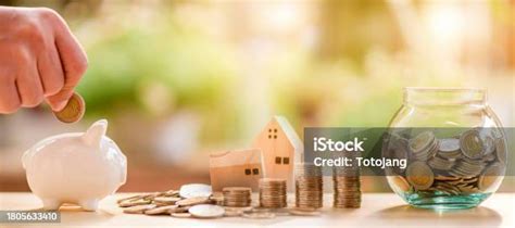 누군가 손에 흰 돼지 저금통을 들고 나무 탁자 위에 동전을 쌓고 있습니다 집을 사기 위한 나무로 되는 저축 낱말 돈 경제에 대한