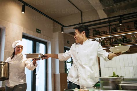 Happy Male Chef Passing Tray To Female Coworker With Salad Bowl In Hand At Commercial Kitchen