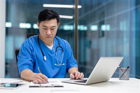 현대적인 사무실의 책상에서 일하는 아시아 남자 의사는 파란색 스크럽을 입고 노트북을 사용하고 환자 상담을 위해 메모를 작성합니다 건강관리와 의술에 대한 스톡 사진 및 기타