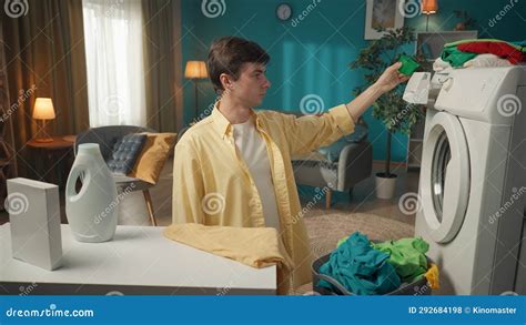 Dark Haired Man Sitting Next To A Washing Machine Pouring A Liquid Detergent Into The Dispenser