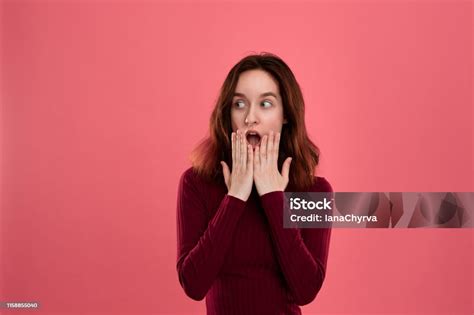 깜짝 놀란 젊은 아가씨는 충격의 감정을 표현하는 입 가까이에서 손을 들고 어두운 분홍색 배경 위에 고립 되어 서 있습니다 갈색