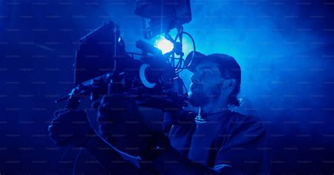 캐주얼웨어를 입은 젊은 남자가 비디오 카메라를 들고 푸른 빛으로 불을 밝히고 연기로 가득 찬 스튜디오에서 광고를 촬영하고 있다