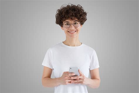 회색 배경에 고립되어 서서 스마트 폰을 들고 귀여운 미소로 카메라를 바라 보는 여성 T 셔츠에 대한 스톡 사진 및 기타 이미지