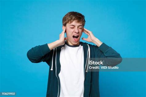스튜디오에서 파란색 배경에 고립 된 손으로 귀를 덮고 눈을 닫힌 유지 캐주얼 복장에 젊은 남자의 초상화 사람들은 진심으로 감정 라이프 스타일 개념입니다 복사 공간을 모의