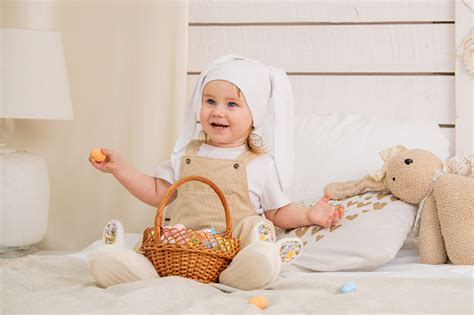 토끼 귀 모자를 쓴 사랑스러운 유아 소녀가 화려한 부활절 달걀로 가득 찬 고리 버들 바구니와 함께 아늑한 침대에 앉아 있습니다 계절에 대한 스톡 사진 및 기타 이미지