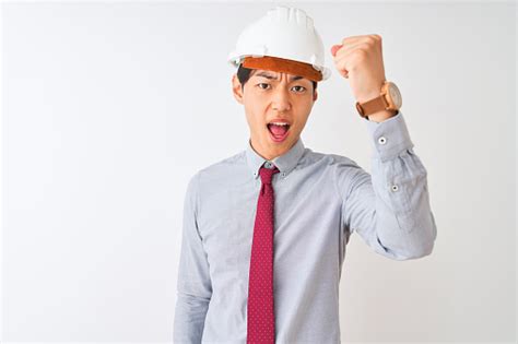 넥타이와 헬멧을 쓰고 고립된 흰색 배경 위에 서있는 중국 건축가 남자는 분노와 함께 외치면서 좌절과 분노를 제기 주먹을 제기 분노와 공격적인 개념 감정에 대한 스톡 사진 및
