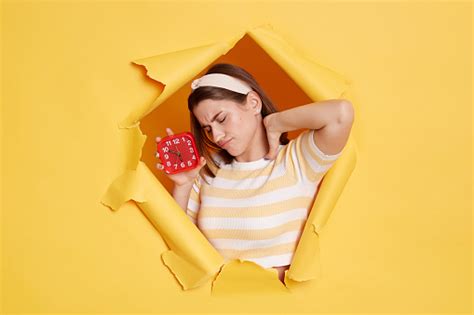 줄무늬 티셔츠와 헤어 밴드를 착용하고 노란 벽의 종이 구멍을 뚫고 알람 시계를 보여주고 목에 통증을 느끼는 건강에 해로운 지친