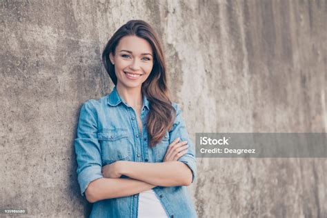 귀여운 갈색 머리 아가씨 콘크리트 벽을 야외에서 웃 고 캐주얼 데님 청바지 복장에서 교차 팔 기울고 있다 갈색 머리에 대한 스톡 사진 및 기타 이미지 Istock