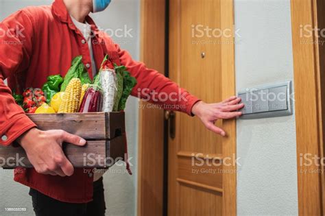 현관에 음식과 종이 봉투를 들고 보호 마스크에 배달 남자 택배는 문에 서서 고객의 초인종을 울립니다 그는 신선한 채소와 과일 한